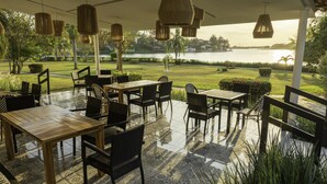 Uma área de refeições ao ar livre com mesas e cadeiras, rodeada por vegetação e vista para um lago.