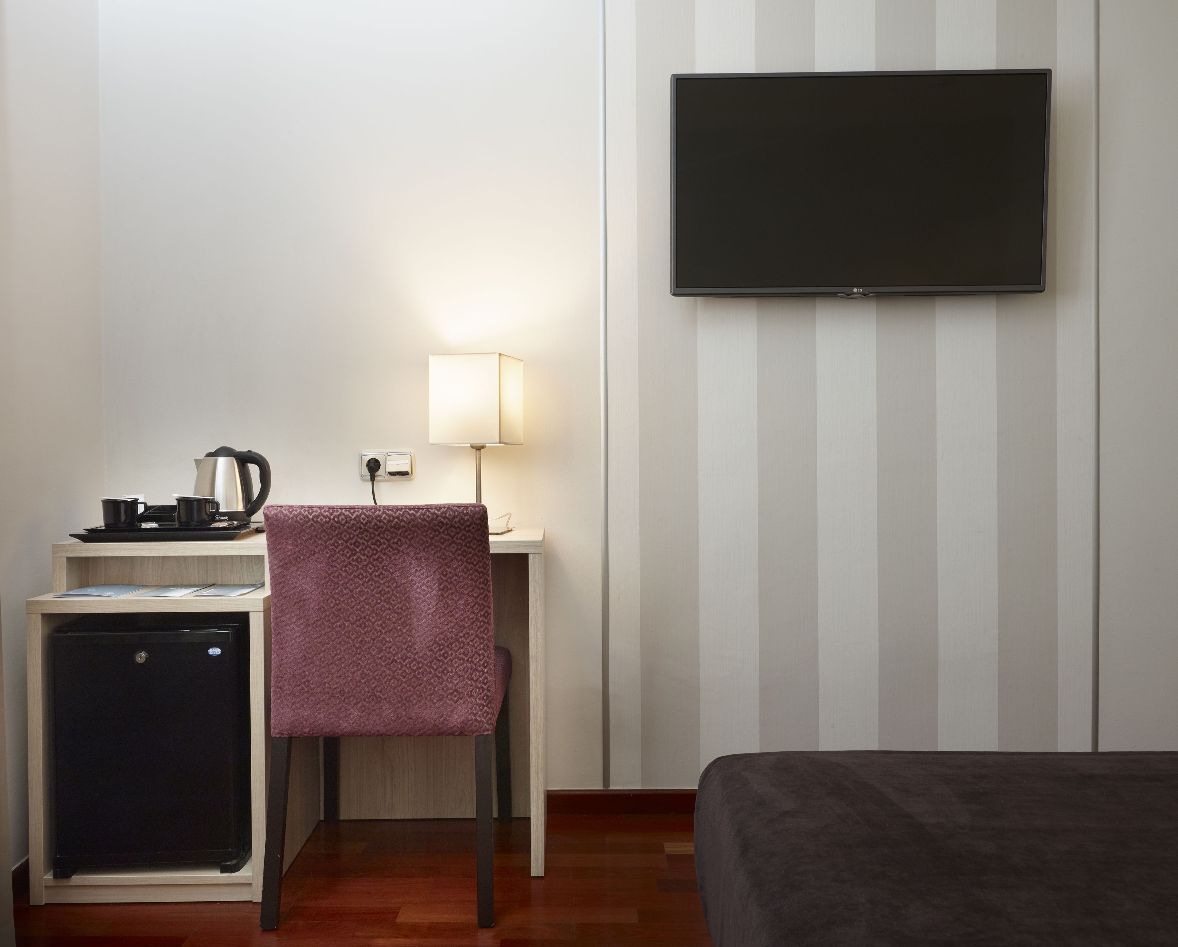Quarto de hotel com cama, escrivaninha com chaleira, luminária e TV na parede.