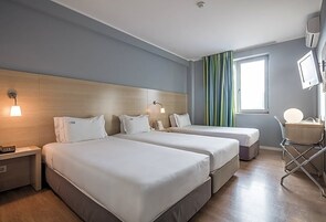 Quarto de hotel com duas camas, uma escrivaninha, uma cadeira, televisão e janela com cortinas.
