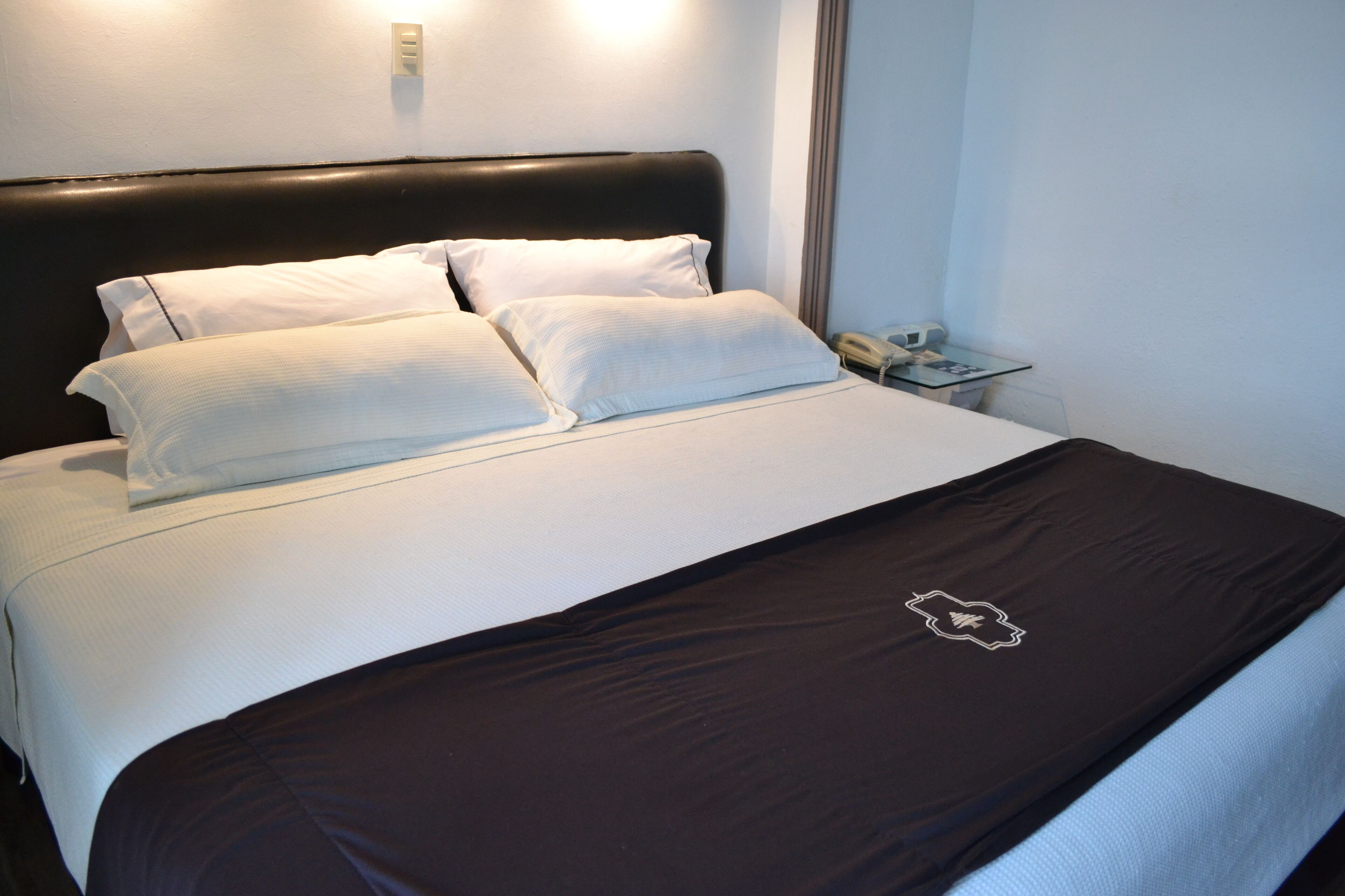 Cama bem arrumada com lençóis brancos e um cobertor marrom escuro, um telefone de mesa e um luminário fixado na parede.
