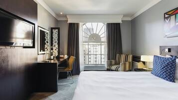 Quarto de hotel com cama, escrivaninha, cadeira e uma janela grande com cortinas.