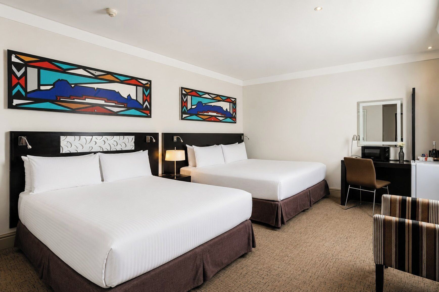Quarto de hotel com duas camas, uma escrivaninha, uma cadeira e dois quadros abstratos na parede.