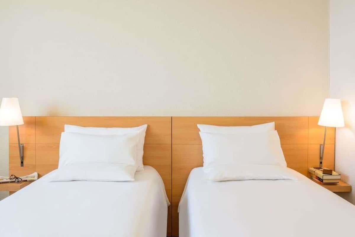 Quarto de hotel com duas camas de solteiro, cabeceiras de madeira, luminárias de cabeceira e mesa de cabeceira com um livro.