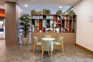 Uma área de recepção com um balcão de madeira, duas cadeiras, uma grande planta em vaso e uma estante com diversos objetos decorativos.