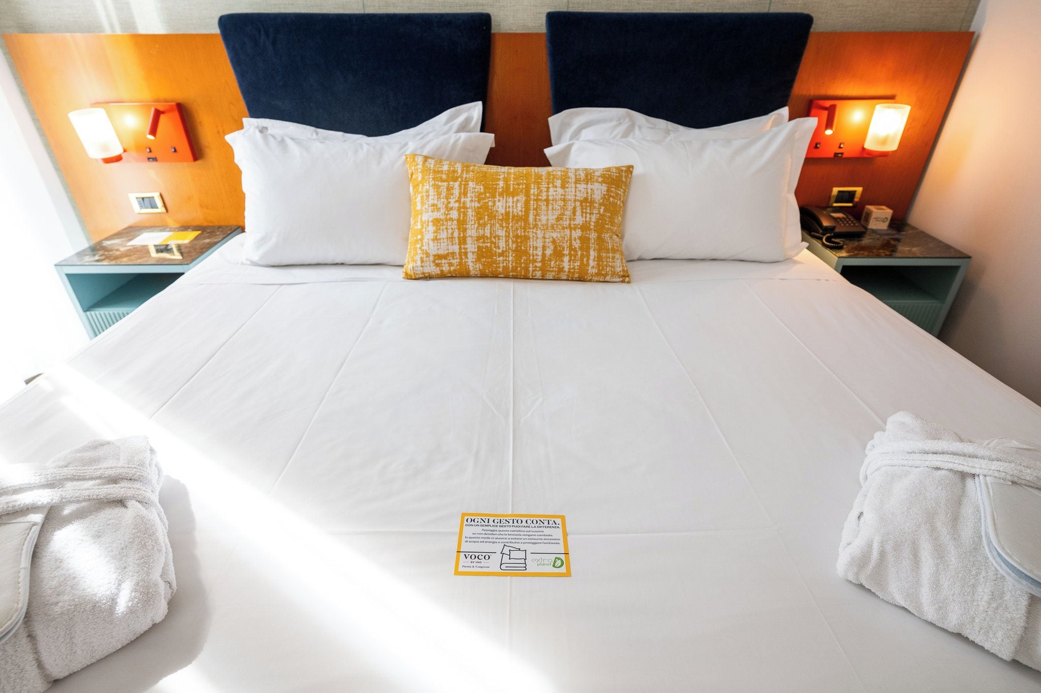 Quarto de hotel com cama, duas almofadas, mesa de cabeceira, luminária e toalha.