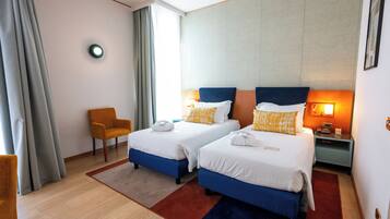 Quarto de hotel com duas camas, uma cadeira, uma mesa de cabeceira e uma janela com cortinas.