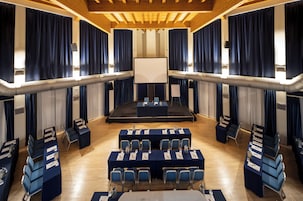 Uma sala de conferências com palco, fileiras de mesas e cadeiras, e uma tela grande.