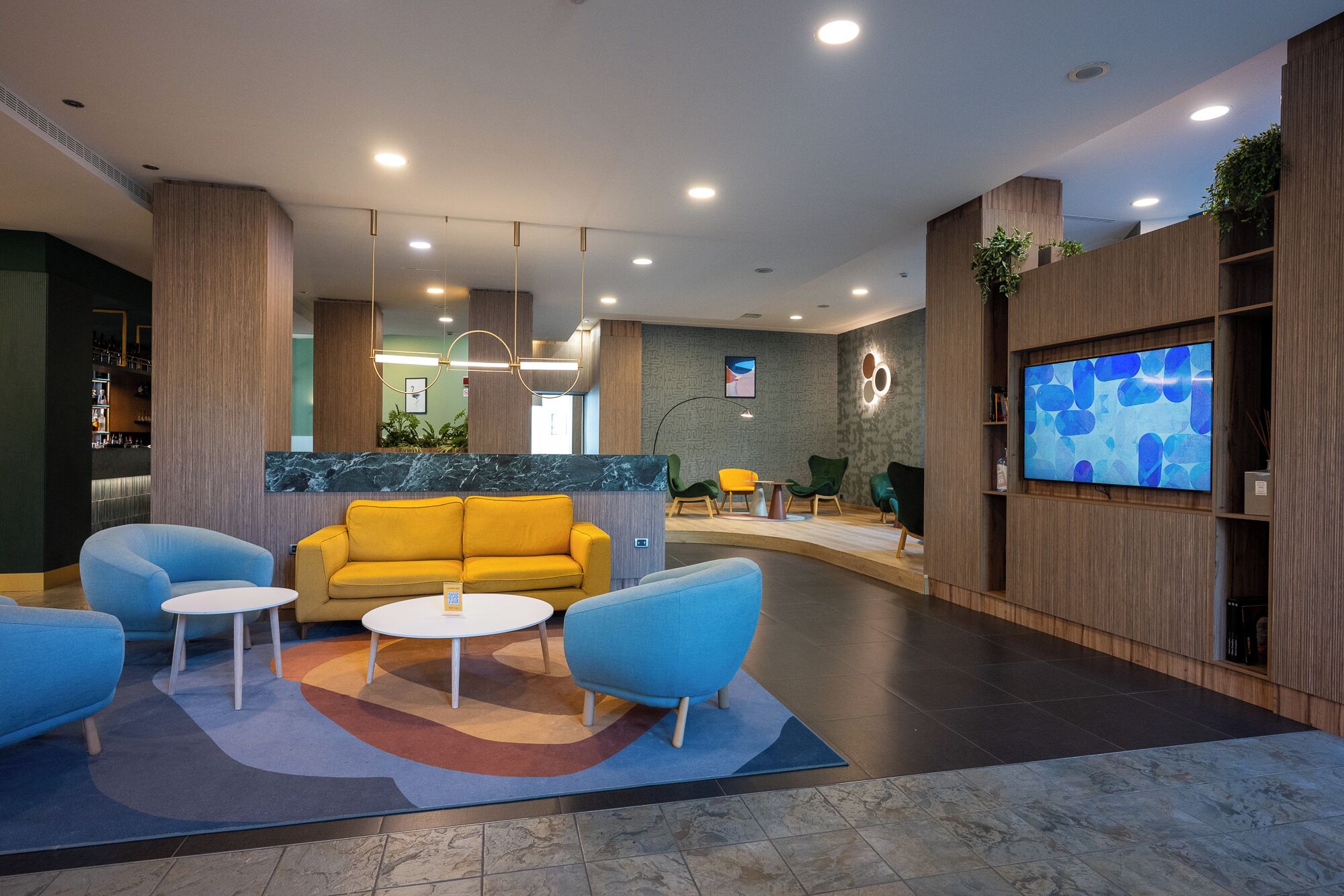 Um hall moderno com um sofá amarelo, poltronas azuis, uma mesa redonda e uma televisão na parede.