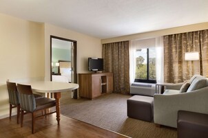 Quarto de hotel com mesa de jantar, duas cadeiras, um sofá, uma televisão em um móvel de madeira e uma janela grande com cortinas.