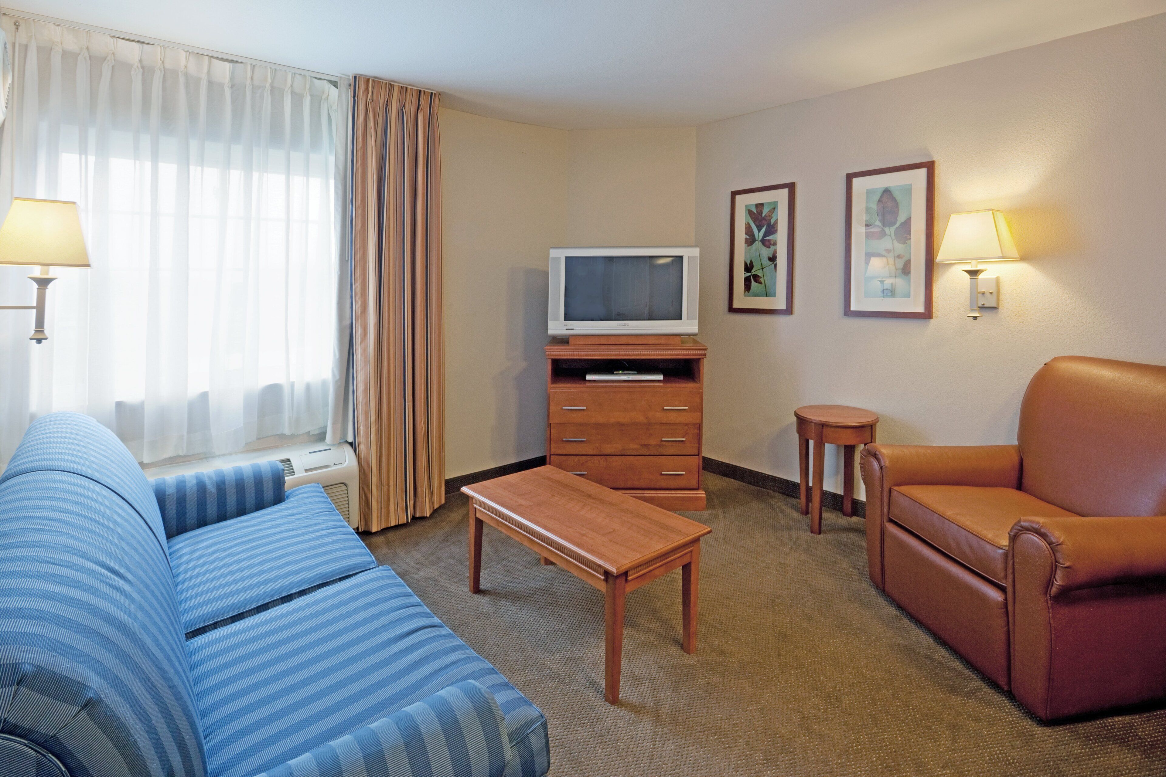 Quarto de hotel com sofá, poltrona, televisão, mesa pequena e duas luminárias.