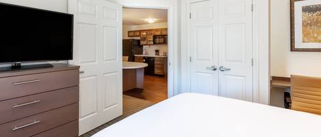 Quarto de hotel com cama, televisão, penteadeira e vista para a cozinha e sala de jantar.