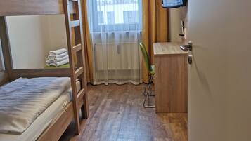 Quarto com beliche, piso de madeira, escrivaninha com cadeira, televisão e janela com cortinas.