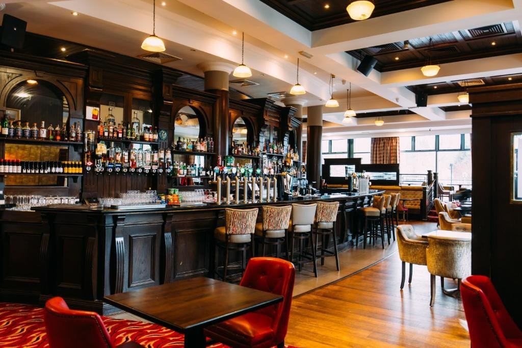 Uma área de bar com balcão de madeira, diversas opções de assento incluindo banquetas e mesas, e variedade de garrafas e copos nas prateleiras.