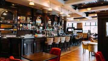 Uma área de bar com balcão de madeira, diversas opções de assento incluindo banquetas e mesas, e variedade de garrafas e copos nas prateleiras.