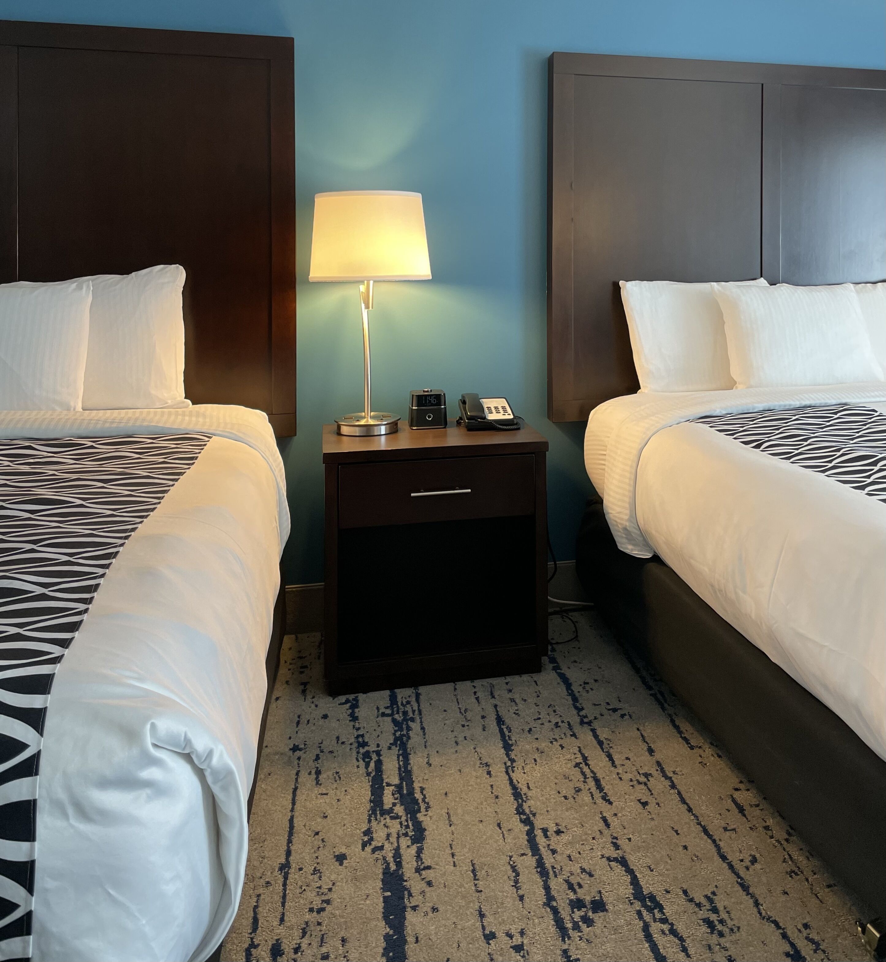 Quarto de hotel com duas camas, uma mesa de cabeceira com abajur, relógio e telefone.