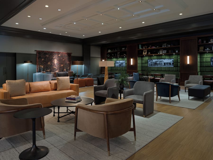Um lobby moderno com sofás e poltronas de couro, e uma mesa de centro. Há livros e um abajur sobre a mesa.