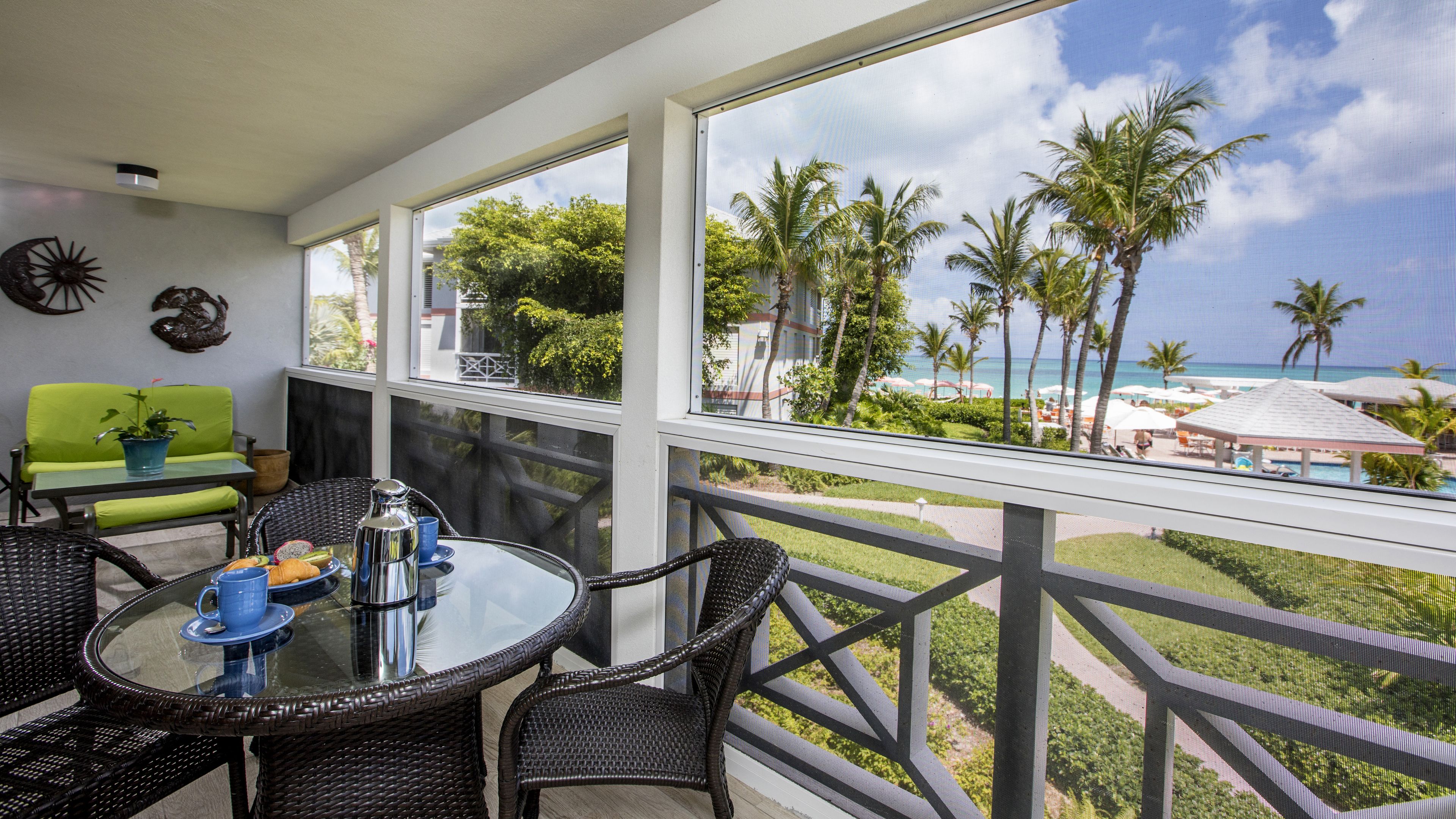 Uma varanda com móveis de vime, uma mesa de vidro e vista para um resort à beira-mar.