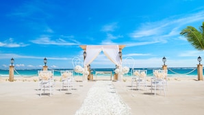 Decoração de casamento na praia, com um corredor branco, cadeiras e um toldo.