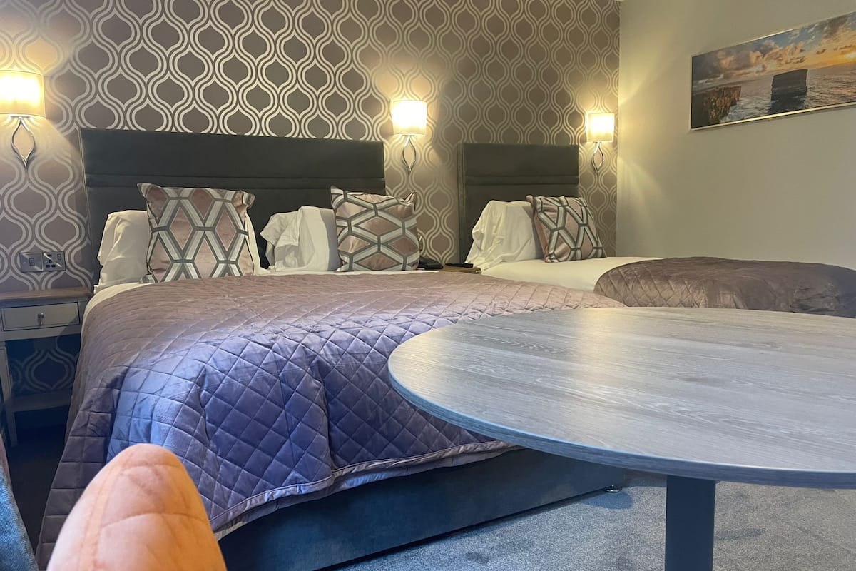 Quarto com cama, duas mesas de cabeceira, uma mesa redonda e uma luminária de parede.