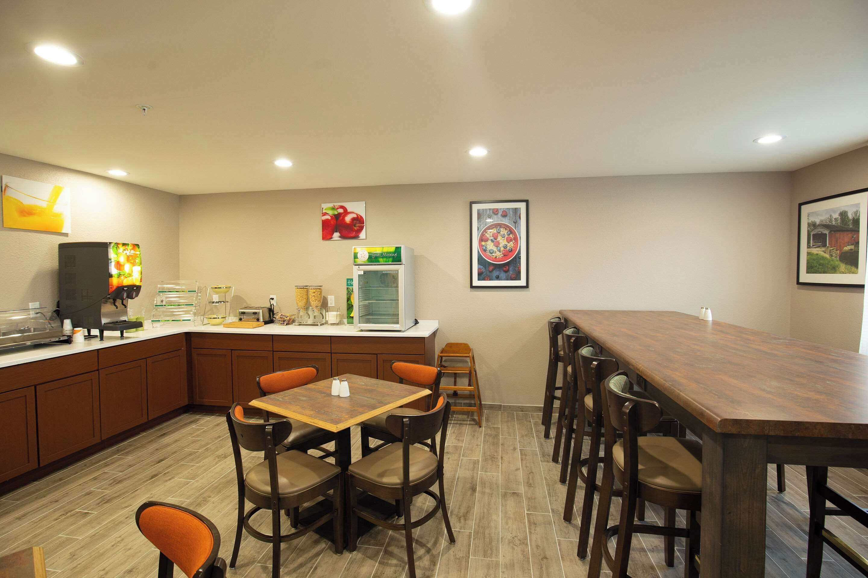 Um restaurante com uma longa mesa de madeira e várias cadeiras, um balcão com produtos alimentícios e um refrigerador.