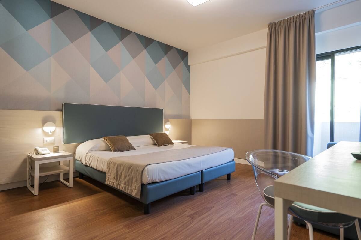 Quarto de hotel moderno com uma cama grande, abajures, uma escrivaninha e vista para o exterior.
