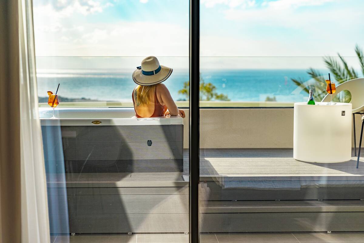 Uma mulher sentada perto de uma janela com vista para o oceano, com uma garrafa e um copo no parapeito.