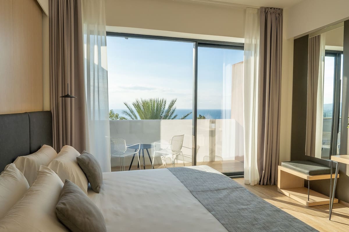 Quarto de hotel moderno com uma cama grande, varanda e vista para o mar, além de um pequeno espaço para sentar.