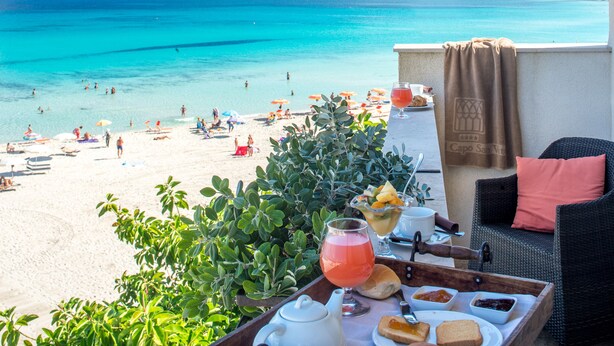 Um terraço com vista para uma praia, uma cadeira de vime, uma mesa com comida e bebidas, e uma toalha pendurada na parede.