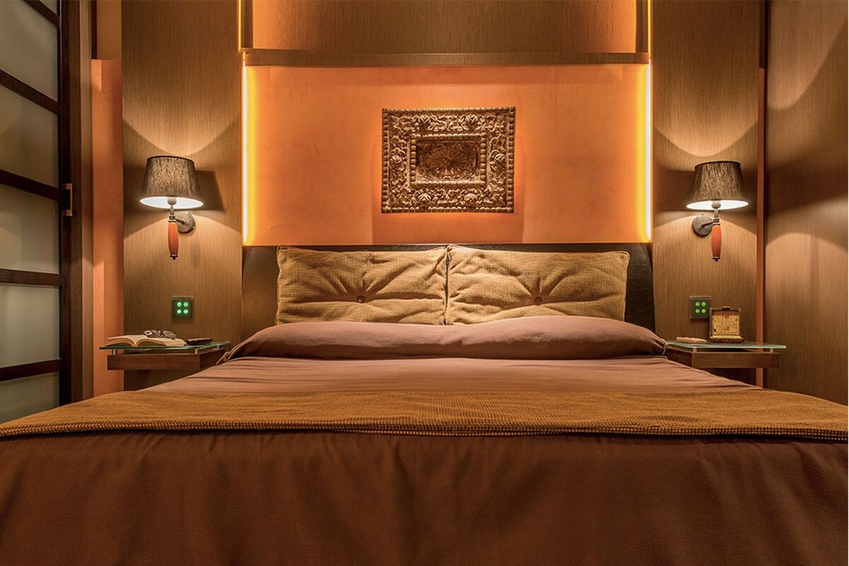 Quarto de hotel com uma cama grande, duas luminárias laterais, um relógio fixado na parede e uma mesinha com um livro e uma luminária.