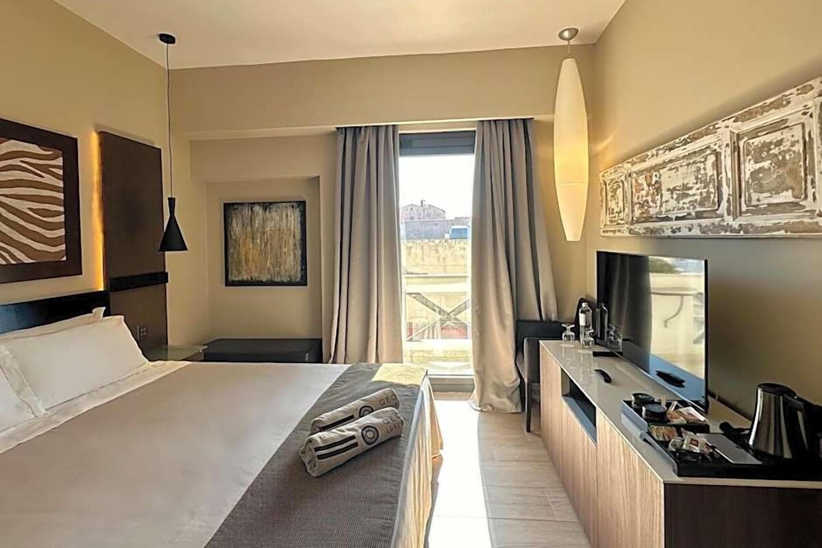 Quarto de hotel com cama, escrivaninha, televisão e janela com cortinas.