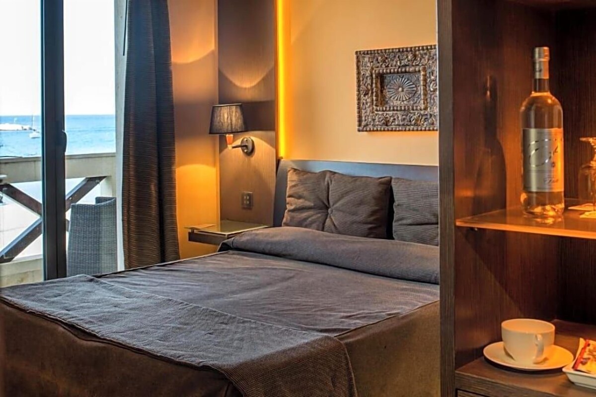 Quarto de hotel com cama, mesa de cabeceira, abajur, relógio de parede, quadro emoldurado e mesa com uma garrafa e uma xícara.