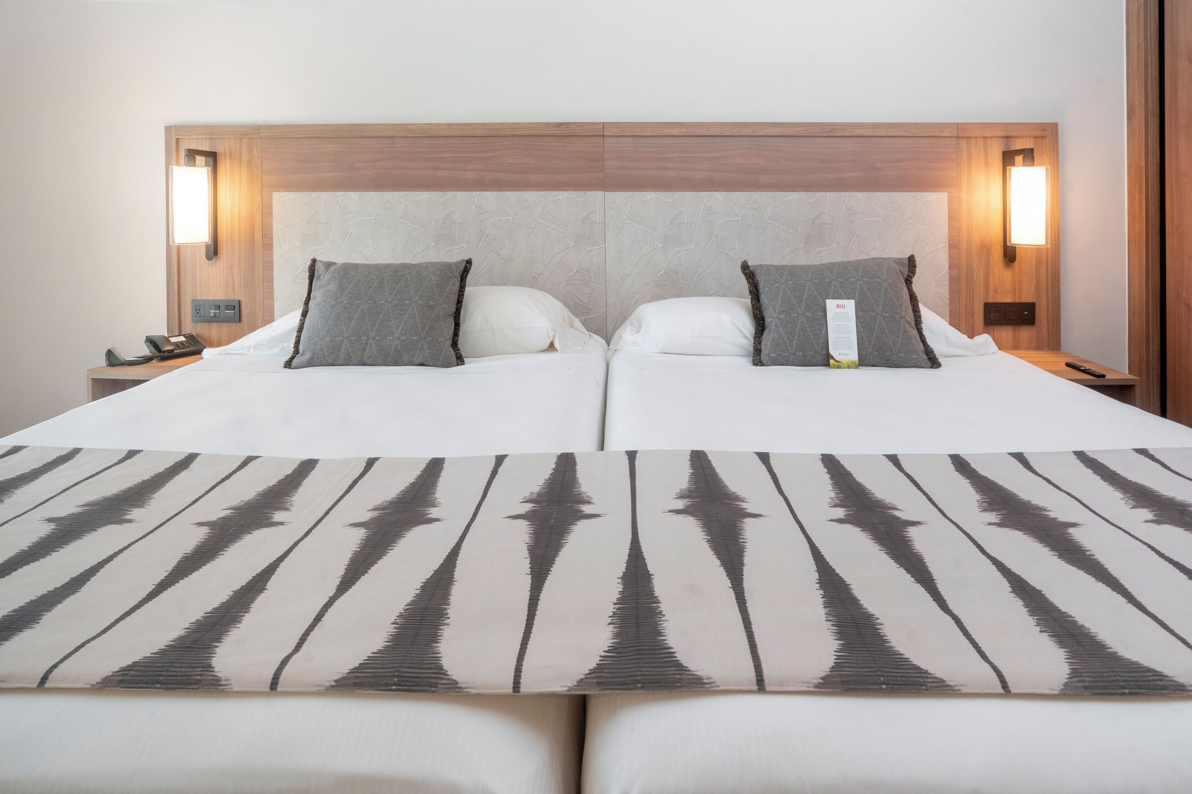 Uma cama de casal com colcha estampada, duas mesas de cabeceira com abajures e uma cabeceira de madeira.