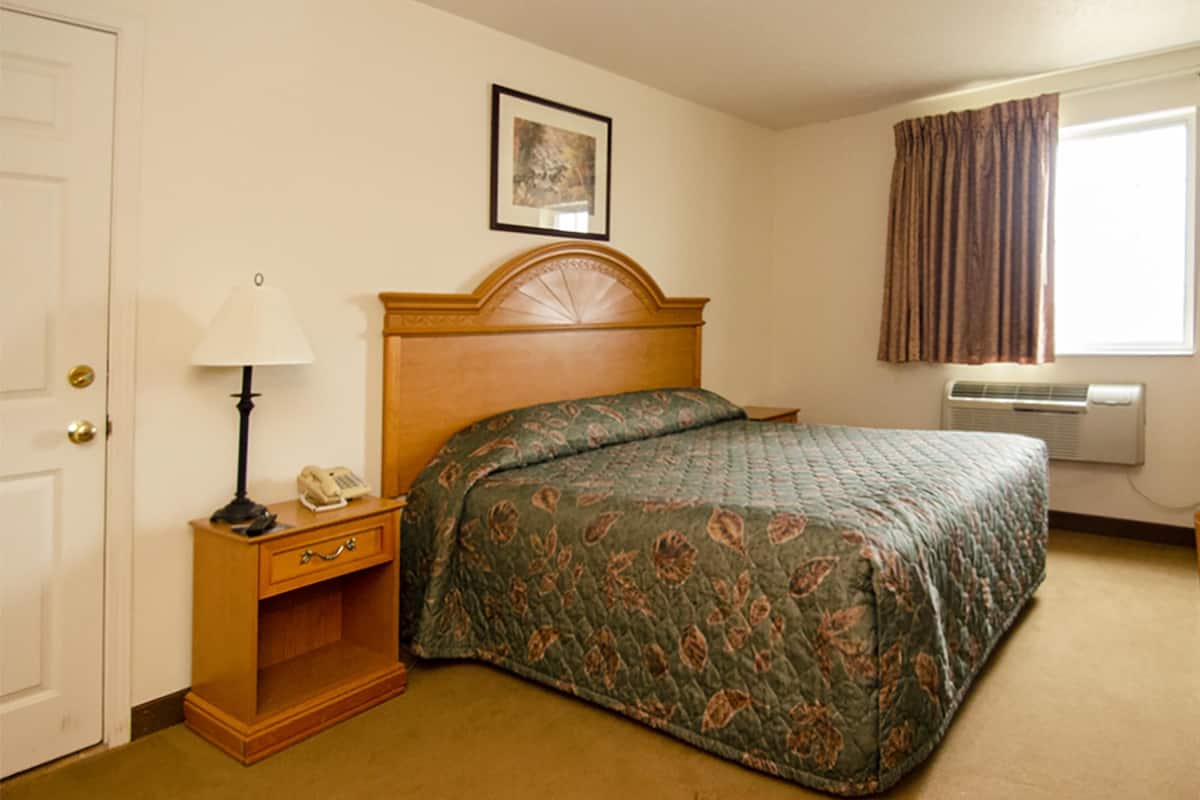 Quarto de hotel com cama, mesa de cabeceira com luminária, telefone, quadro na parede, ar condicionado e janela com cortinas.