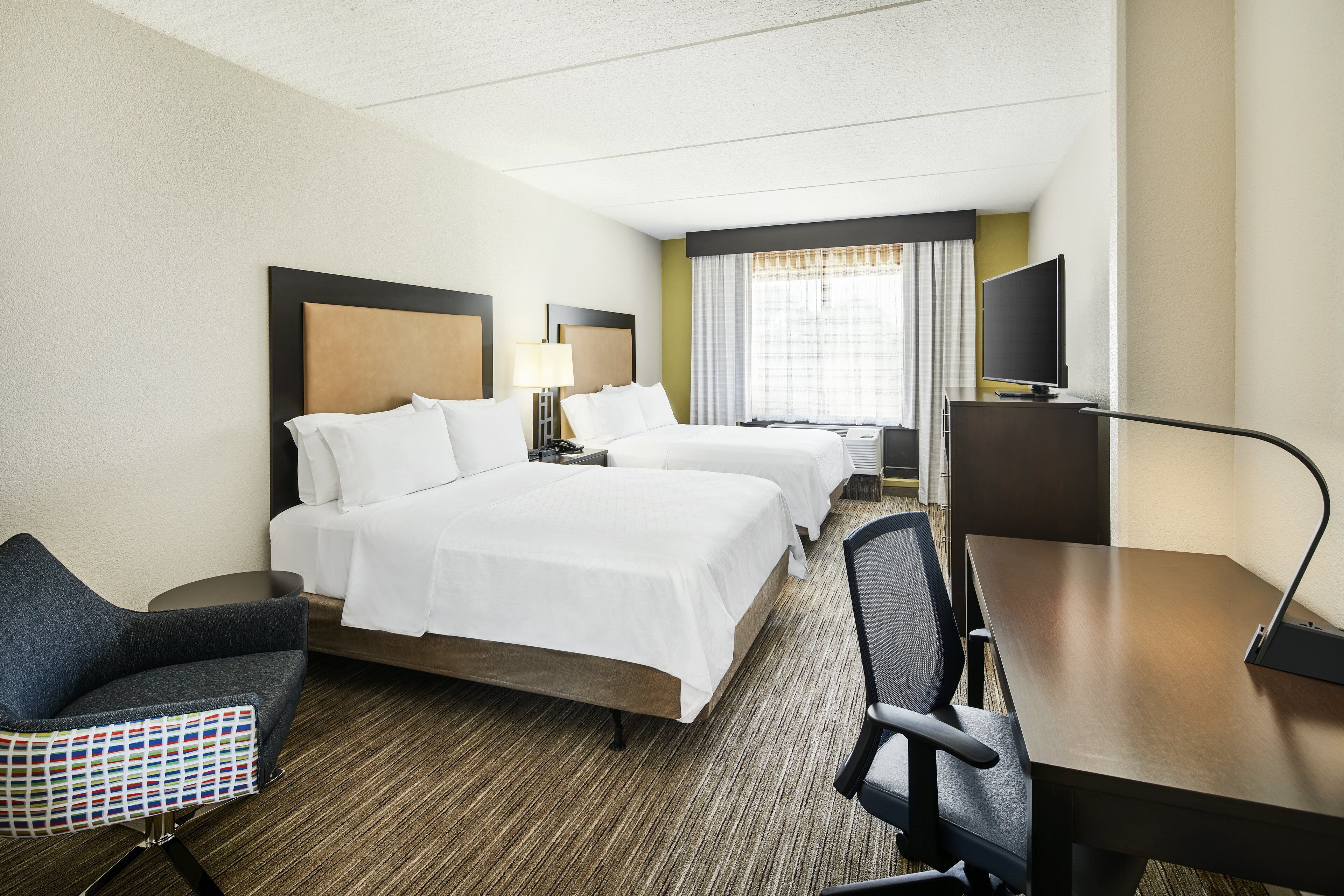 Quarto de hotel com duas camas, uma escrivaninha, uma cadeira, televisão e janela com cortinas.