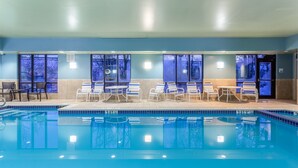 Piscina interna com espreguiçadeiras e mesas, amplas janelas com vista para árvores e piso revestido de azulejos.