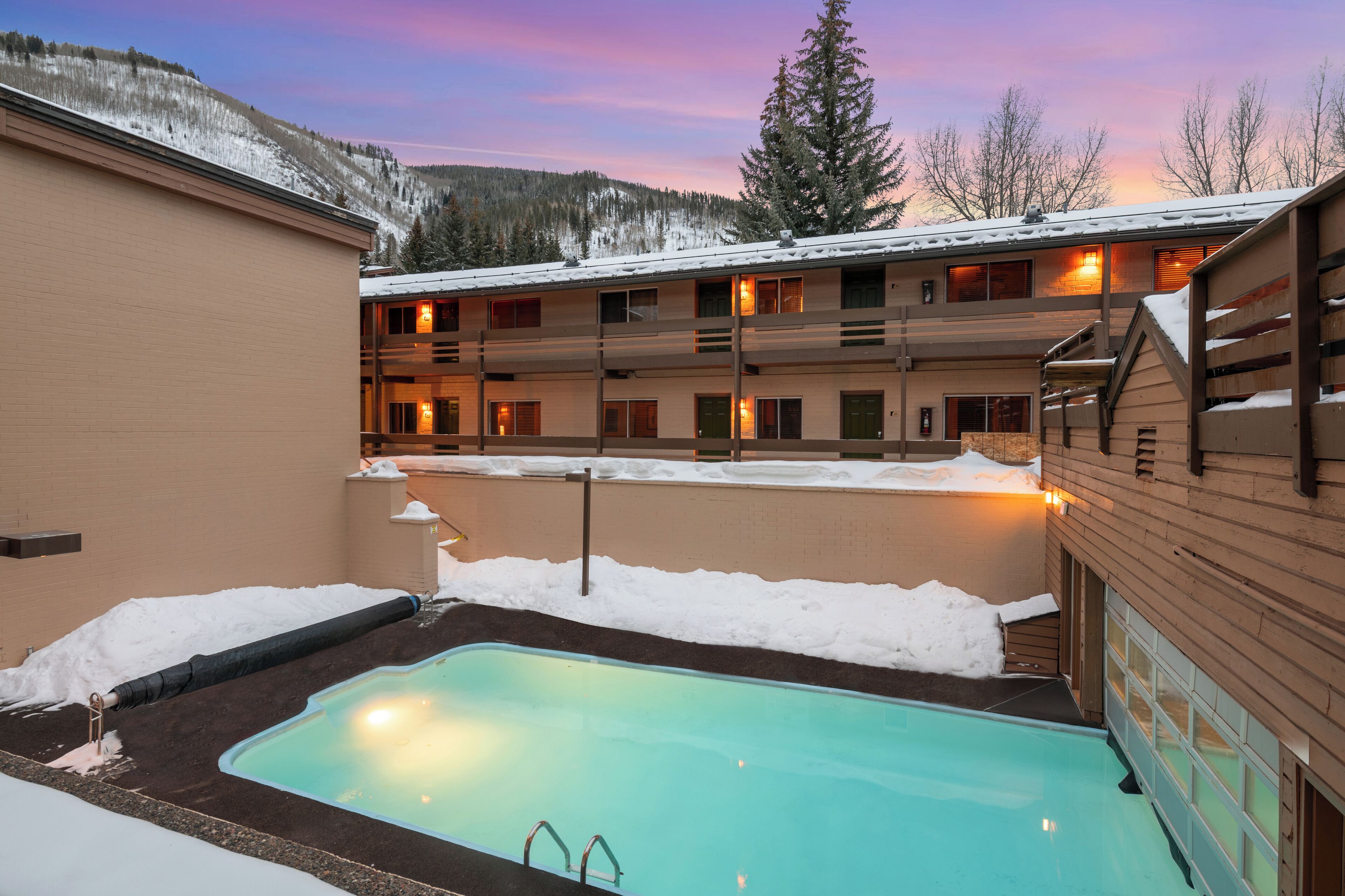 Hotel com piscina rodeada de neve, montanhas ao fundo e um edifício alto com janelas iluminadas.