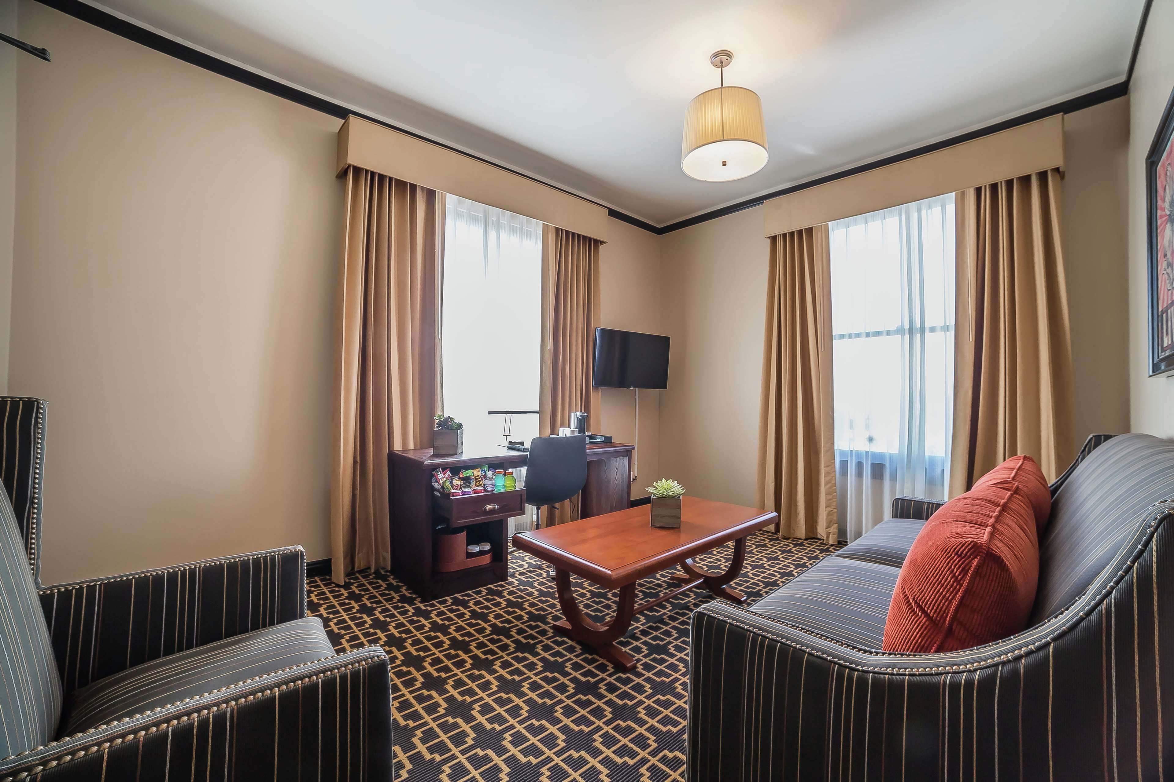 Quarto de hotel com sofá, poltronas, mesa de centro de madeira, escrivaninha e televisão de tela plana, além de carpete estampado.