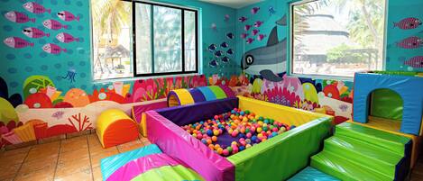 Uma área de recreação com estruturas acolchoadas coloridas e uma piscina de bolinhas, decorada com murais de vida marinha.