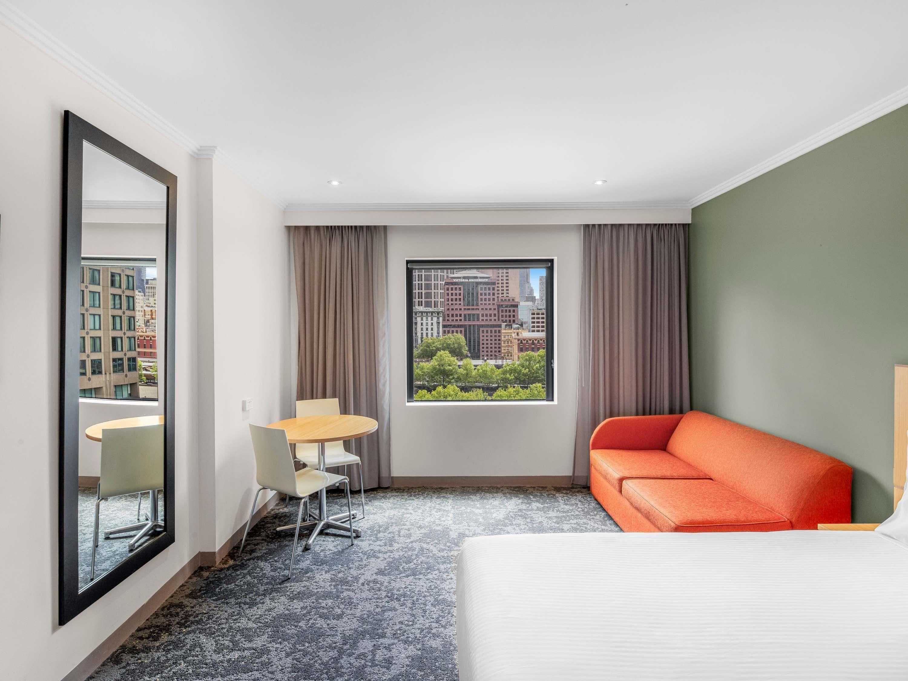 Quarto de hotel com cama, sofá, mesa de jantar com cadeiras, uma grande janela com vista para a cidade e um espelho.