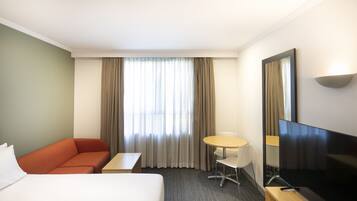 Quarto de hotel com cama, sofá, mesinha, cadeira, televisão e janela com cortinas.