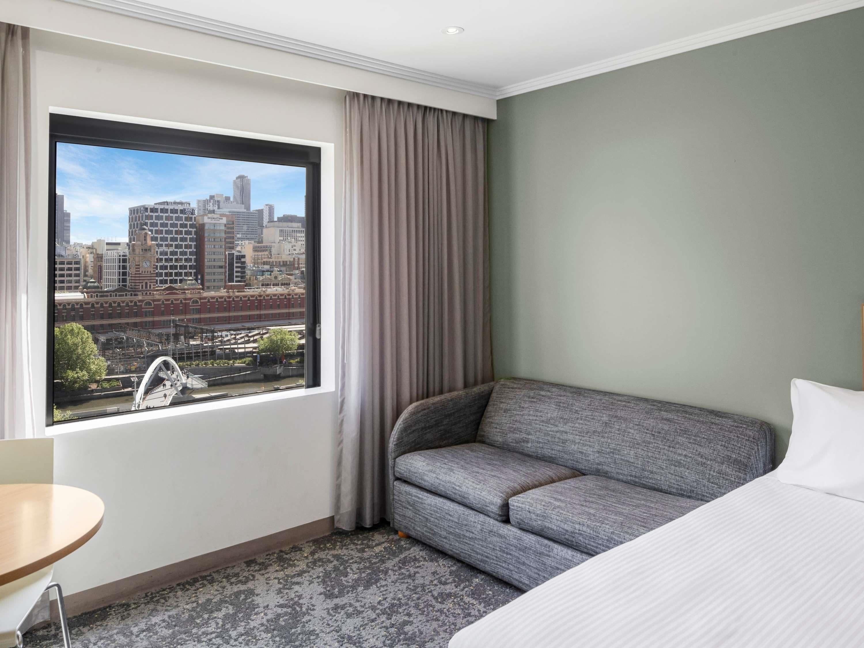 Quarto de hotel com uma janela grande, um sofá cinzento, uma cama e uma pequena mesa.
