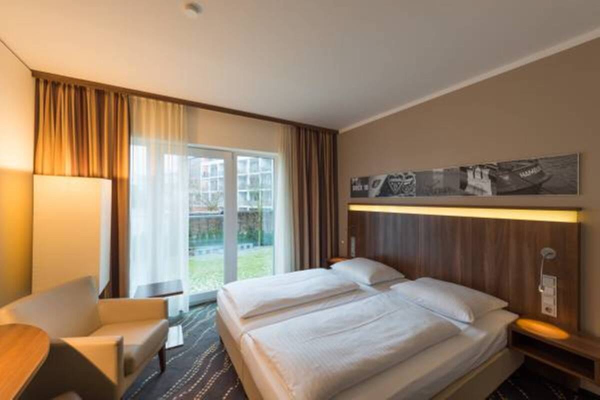 Quarto de hotel com duas camas, uma escrivaninha, uma cadeira, uma janela grande com cortinas e uma obra de arte na parede.