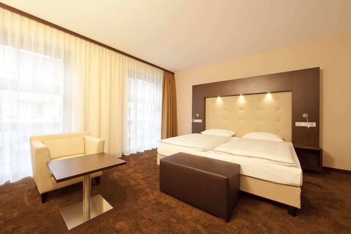 Quarto de hotel com cama de casal, sofá, mesa pequena e uma janela grande com cortinas.