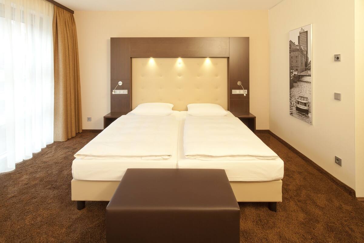 Uma cama de casal com cabeceira, mesas de cabeceira, um banquinho e uma janela grande com cortinas.