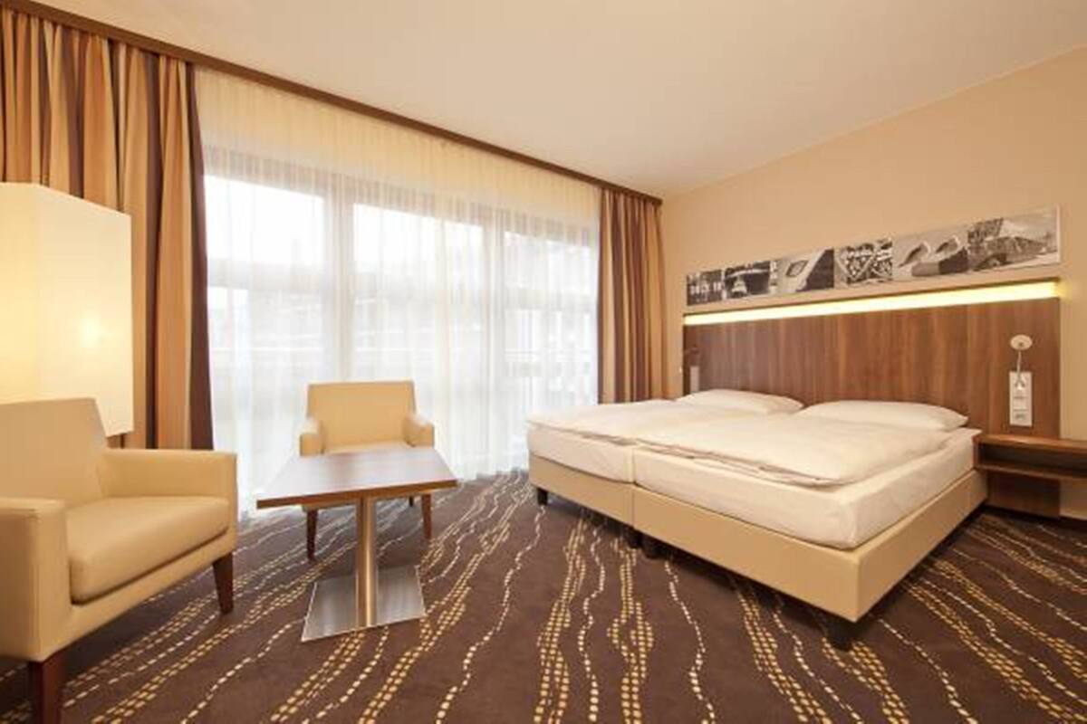 Quarto de hotel com uma cama, uma cadeira, uma mesa e uma janela grande com cortinas.