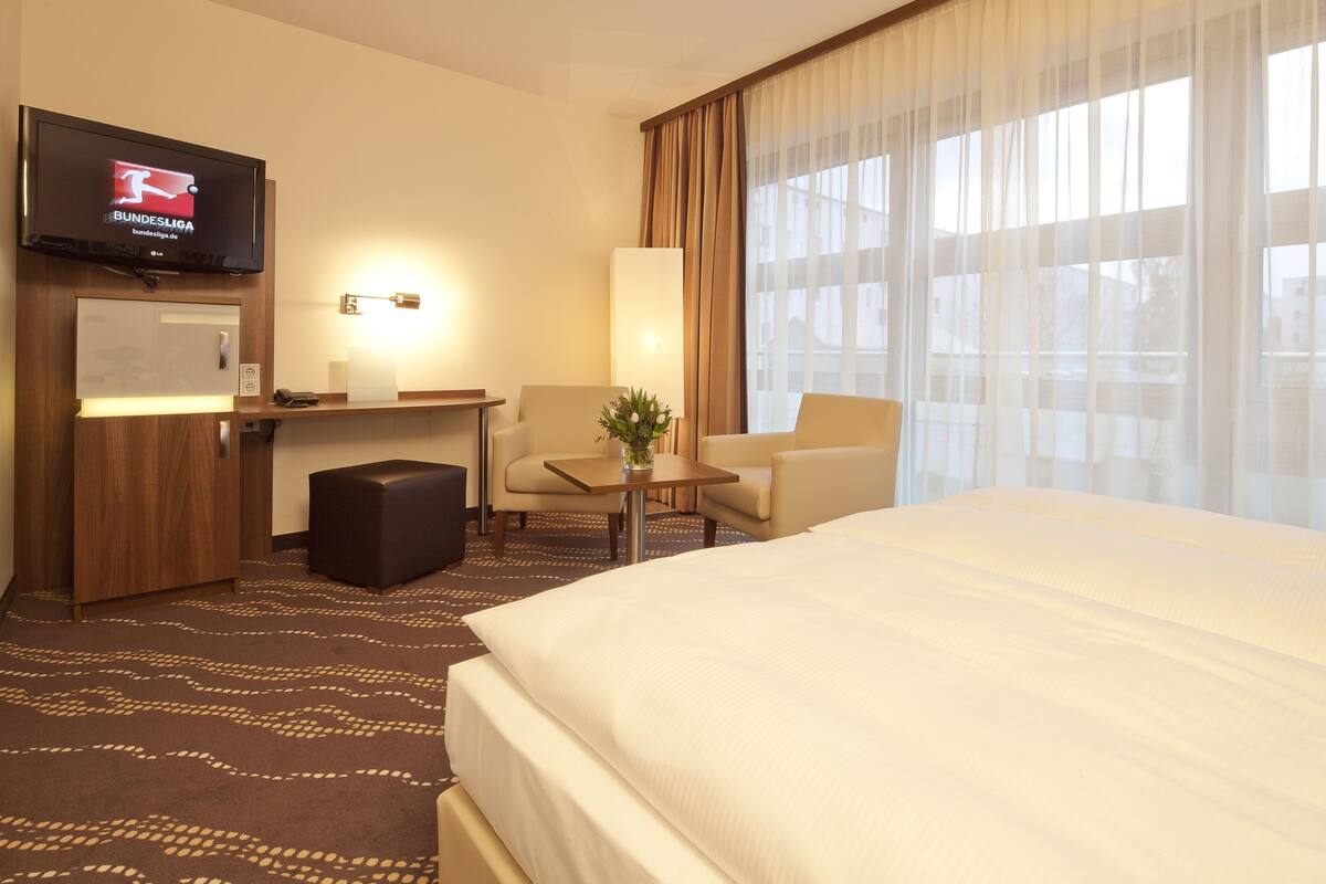Quarto de hotel com cama, escrivaninha, cadeira, televisão e janela com cortinas.