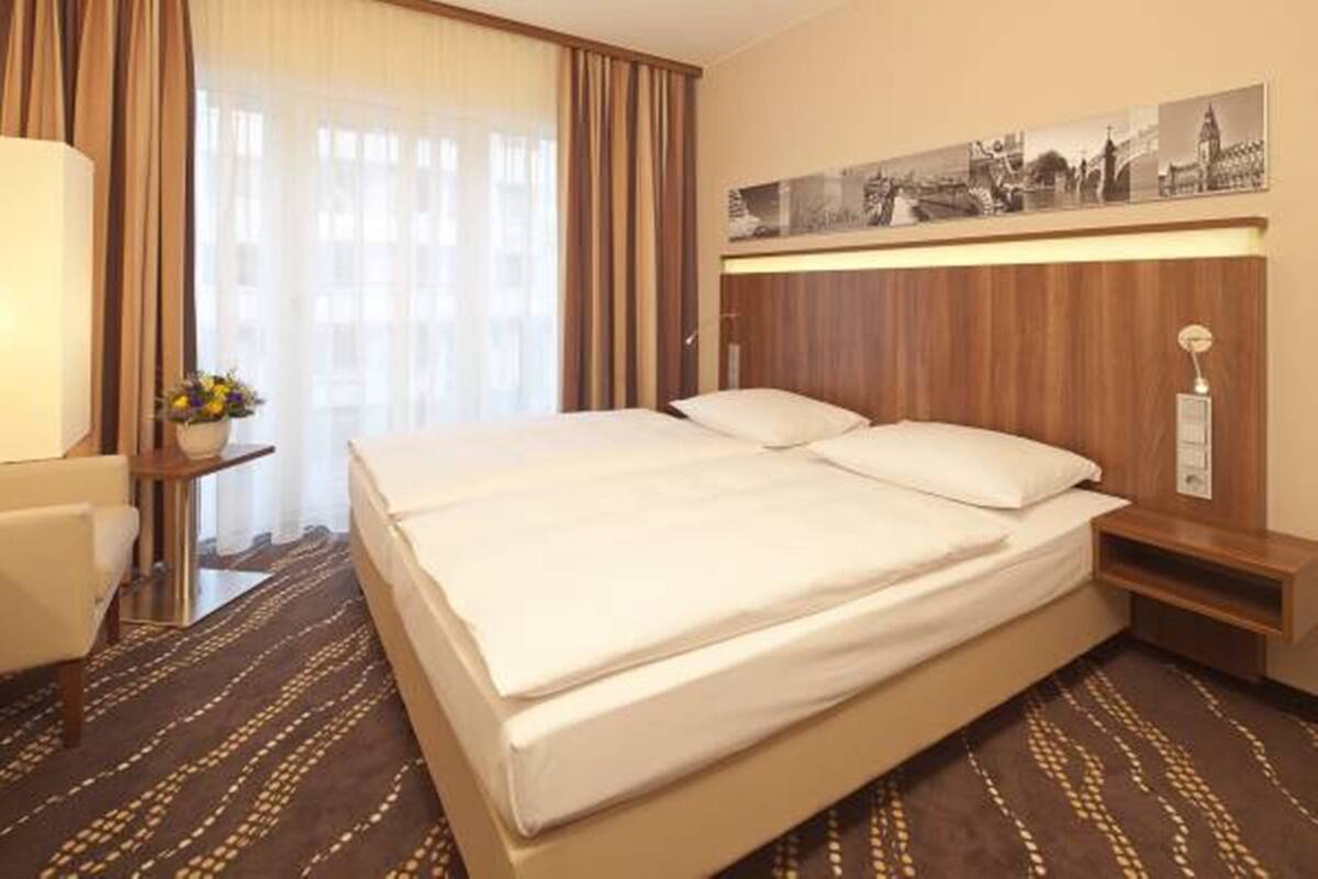 Quarto de hotel com uma cama grande, uma mesa de cabeceira com abajur, uma pequena mesa com um vaso de flores e um quadro na parede.