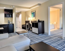 Quarto de hotel com cozinha compacta, mesa, cadeira, sofá e banheiro visível por uma porta aberta.