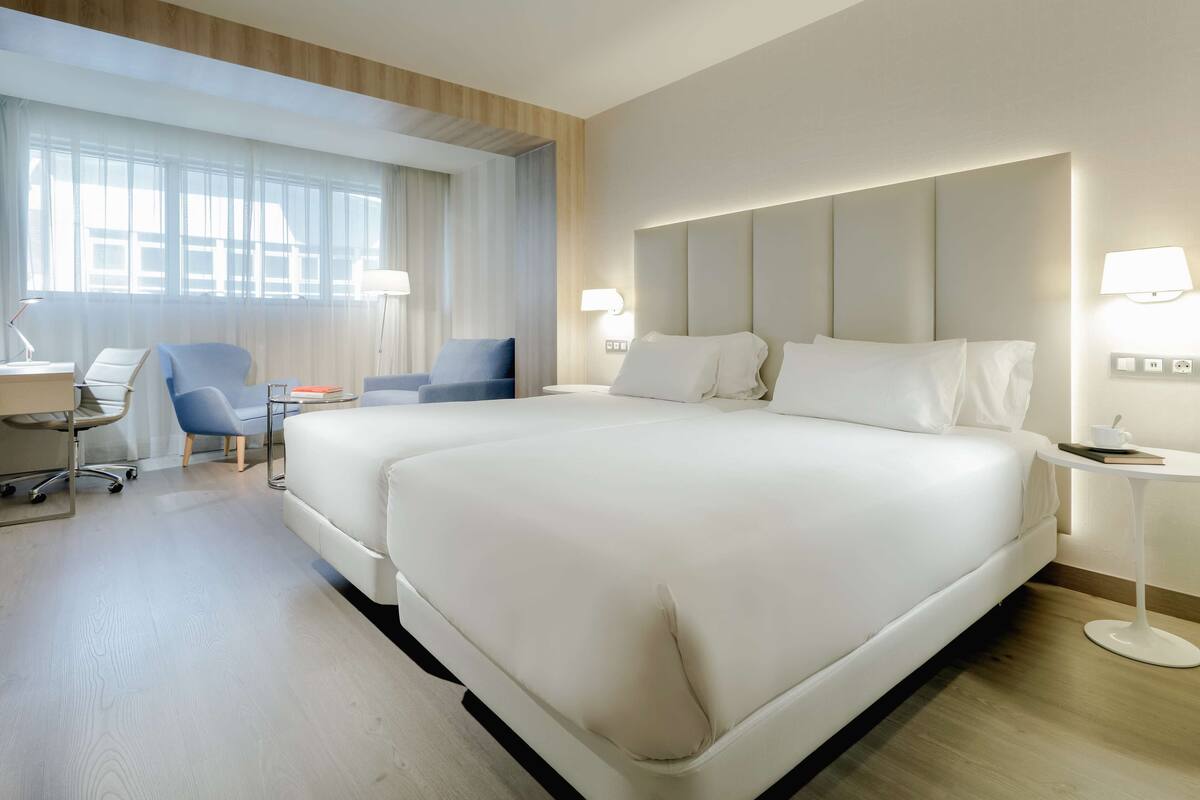 Quarto de hotel moderno com uma cama grande, uma mesa de trabalho com cadeira e uma mesinha de cabeceira com abajur.