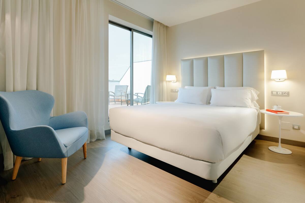 Quarto de hotel com uma cama grande, uma poltrona azul, uma mesa redonda pequena com uma luminária e vista para o exterior através de uma porta deslizante.